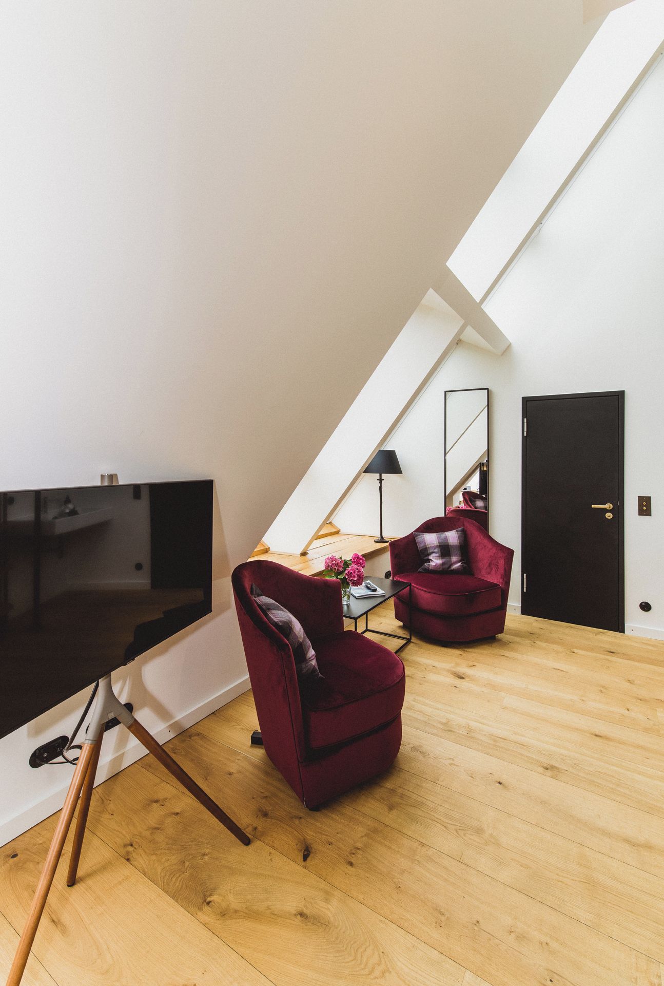 Quarto moderno no estilo loft, com teto inclinado, TV de tela plana, duas poltronas de veludo vermelho, piso de madeira e porta preta.
