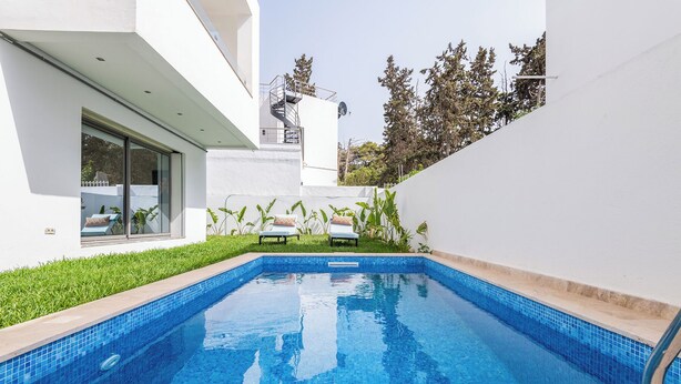 Uma piscina exterior moderna com design de azulejos azuis translúcidos, rodeada por um relvado bem cuidado e um edifício branco com grandes janelas e uma antena parabólica.