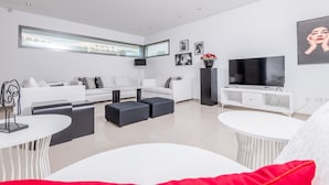 Uma sala de estar moderna com um sofá branco, pufes pretos, uma mesa de centro branca, uma televisão de tela plana e uma grande janela com vista para a cidade.