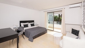 Quarto moderno com cama, escrivaninha, cadeira e sofá. Uma porta de vidro deslizante dá acesso a uma área externa.