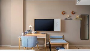 Quarto de hotel moderno com televisão de tela plana na parede, uma escrivaninha de madeira com cadeira, um pequeno banco e uma cama com cabeceira branca.