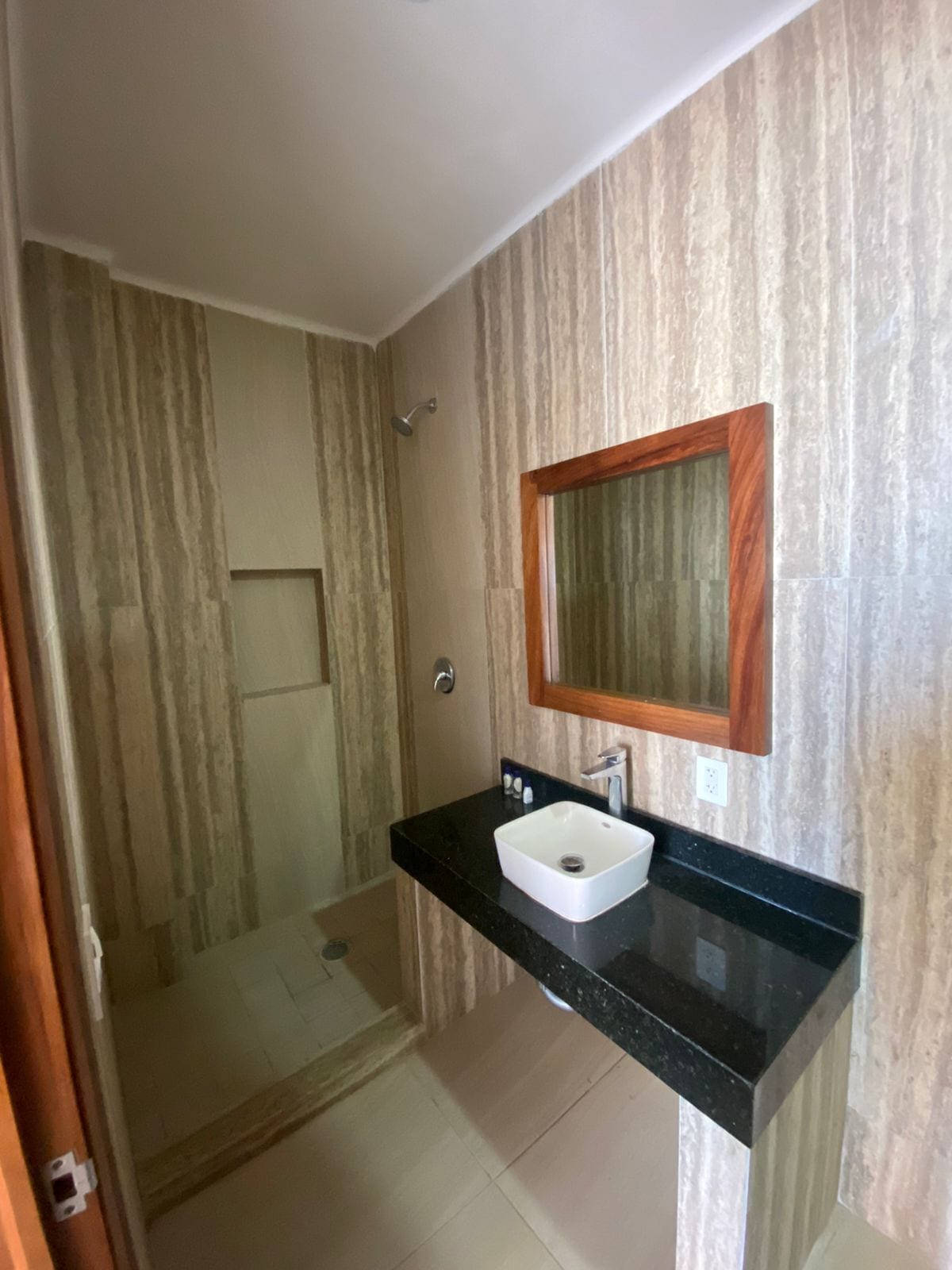 Casa de banho com bancada preta, lavatório branco, espelho com moldura de madeira e zona de duche.