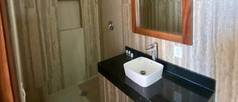 Casa de banho com bancada preta, lavatório branco, espelho com moldura de madeira e zona de duche.