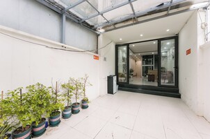 Espaço interno limpo, com piso de azulejos, plantas em vasos e uma porta de vidro que dá para um quarto.