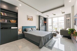 Quarto moderno com cama grande, estantes embutidas, mesa de cabeceira, abajur e vista da cidade através de grandes janelas.