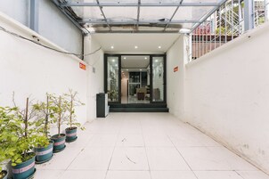 Um corredor limpo e com azulejos, com plantas em vasos à esquerda, que leva a uma entrada com porta de vidro.