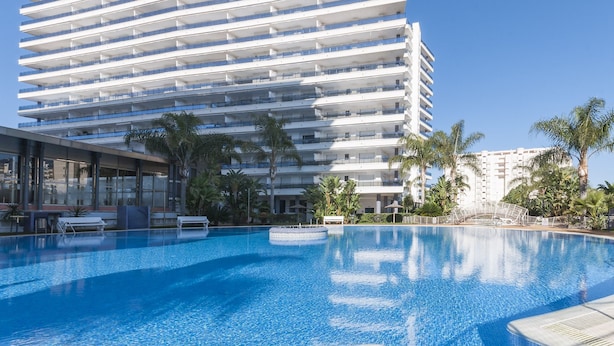 Um hotel moderno com uma piscina grande, palmeiras e um edifício de vários andares com varandas.