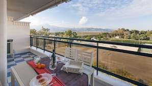 Uma varanda com piso xadrez, uma mesa posta com pratos e copos, uma cadeira e vista para uma paisagem com montanhas e árvores.