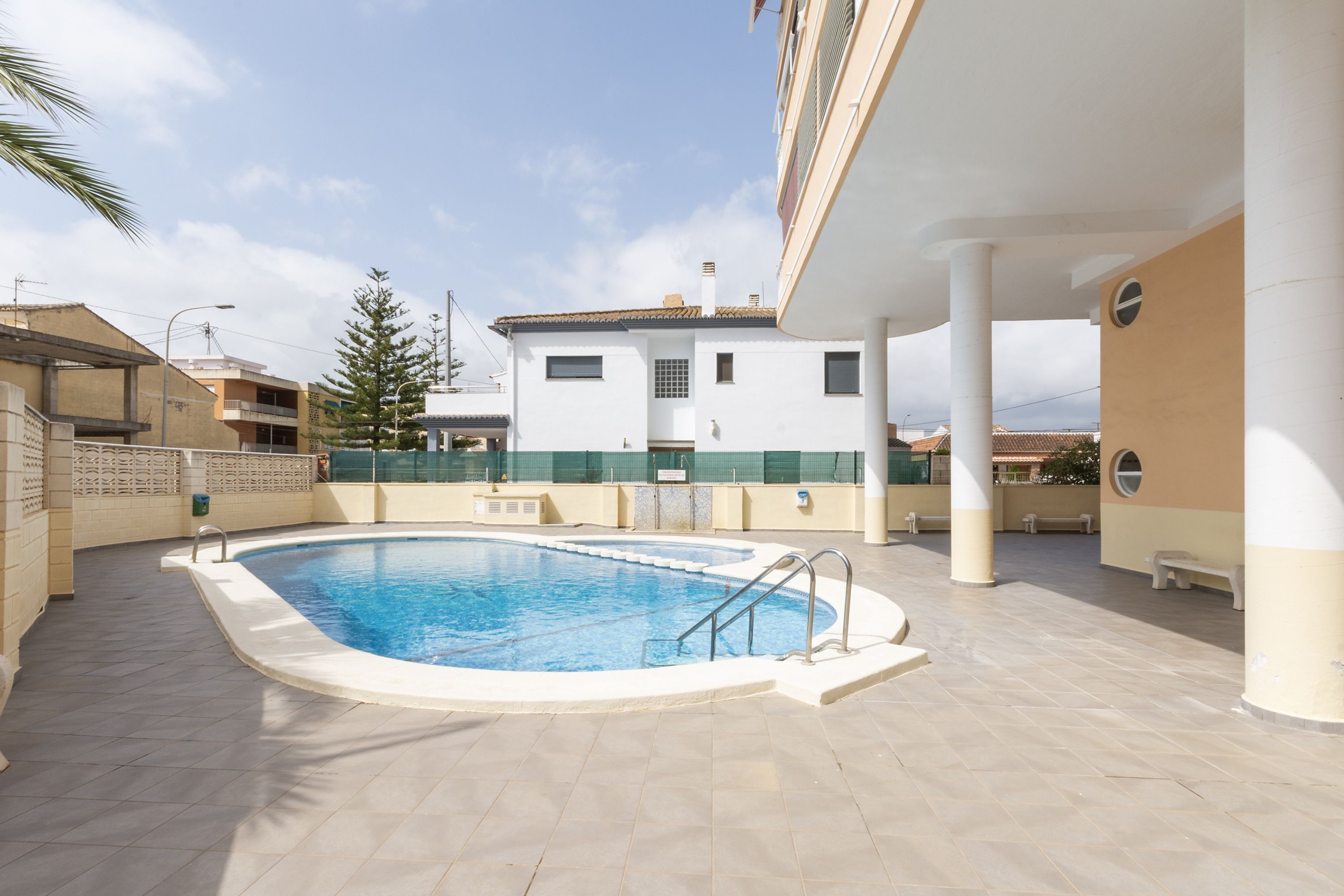 Uma área de piscina com piso ladrilhado, piscina com escada de metal e um prédio com varandas ao fundo.