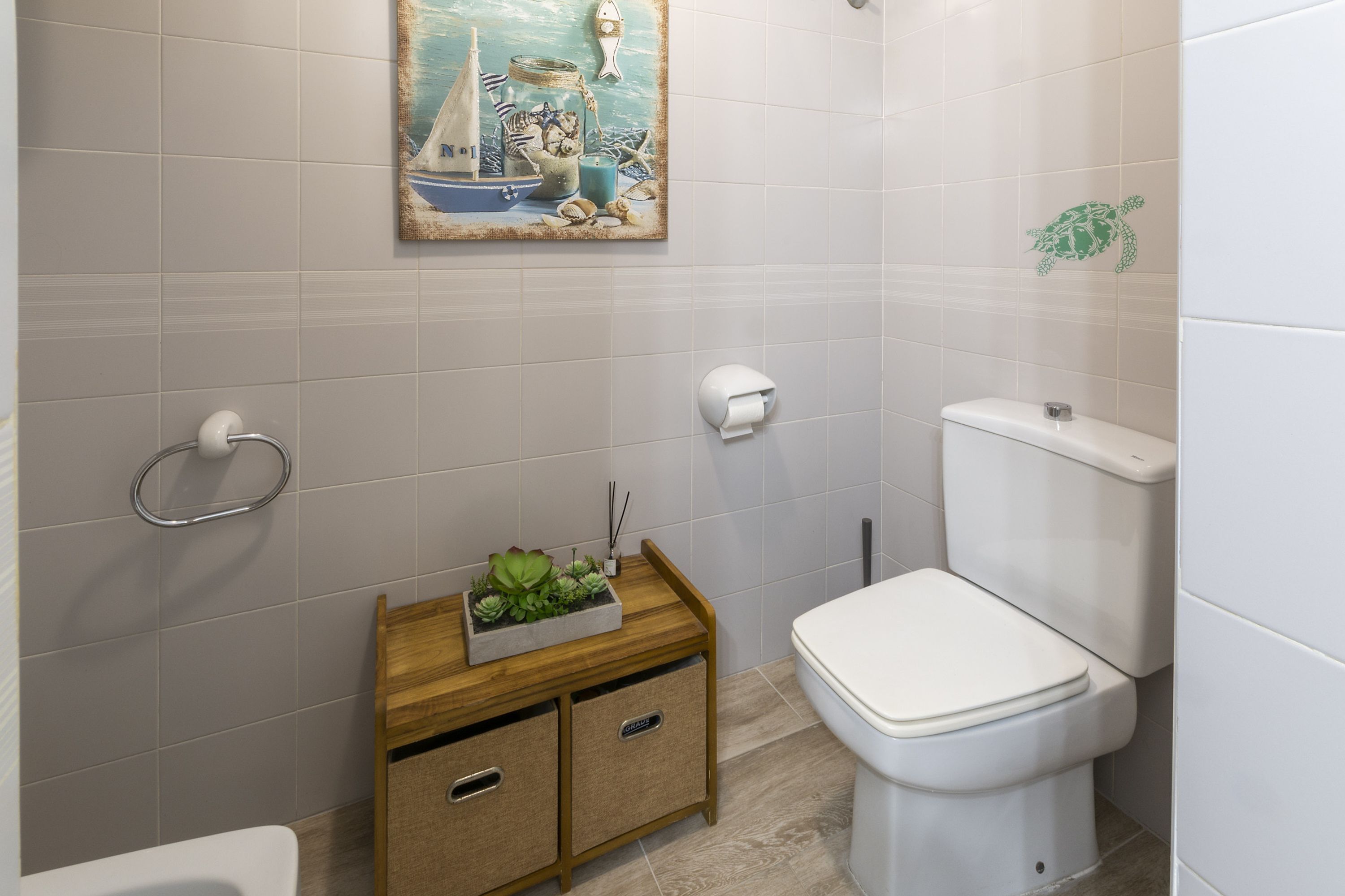 Casa de banho com sanita, um pequeno armário de madeira com uma planta, um porta-toalhas e um suporte de papel higiénico fixado na parede.