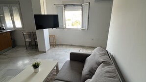 Uma sala de estar moderna com um sofá cinzento, uma mesa de centro branca e uma pequena planta. Há uma televisão na parede e uma janela com persianas.