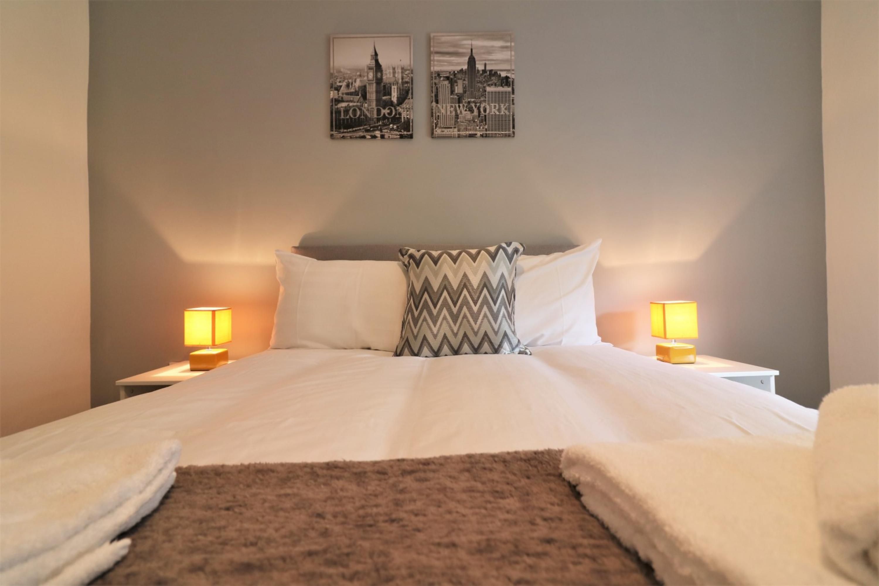 Cama bem arrumada com lençóis brancos, dois travesseiros, um cobertor marrom e duas luminárias de cabeceira.
