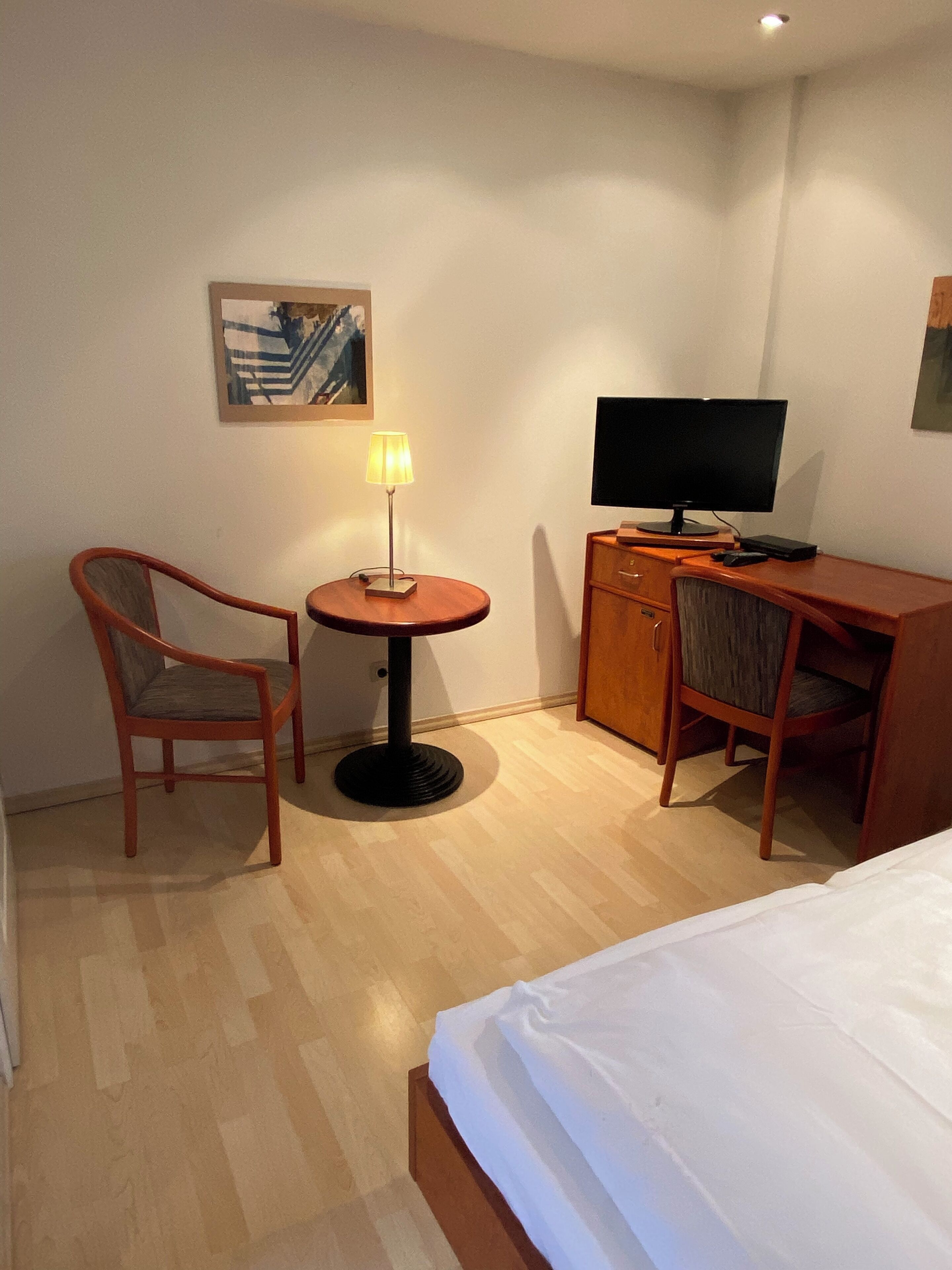 Quarto de hotel com uma cama, uma escrivaninha com televisão, uma cadeira e um abajur.