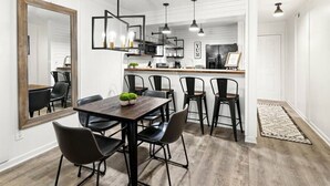 Uma área de refeições moderna com uma mesa de madeira e cadeiras pretas, uma cozinha ao fundo e iluminação pendente.