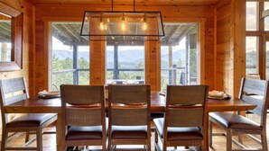 Interior de madeira com uma mesa de jantar e cadeiras, grandes janelas com vista para a montanha e um lustre.