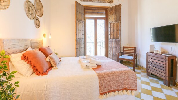 Quarto bem arrumado com cama, mesa de cabeceira, cadeira e televisão.