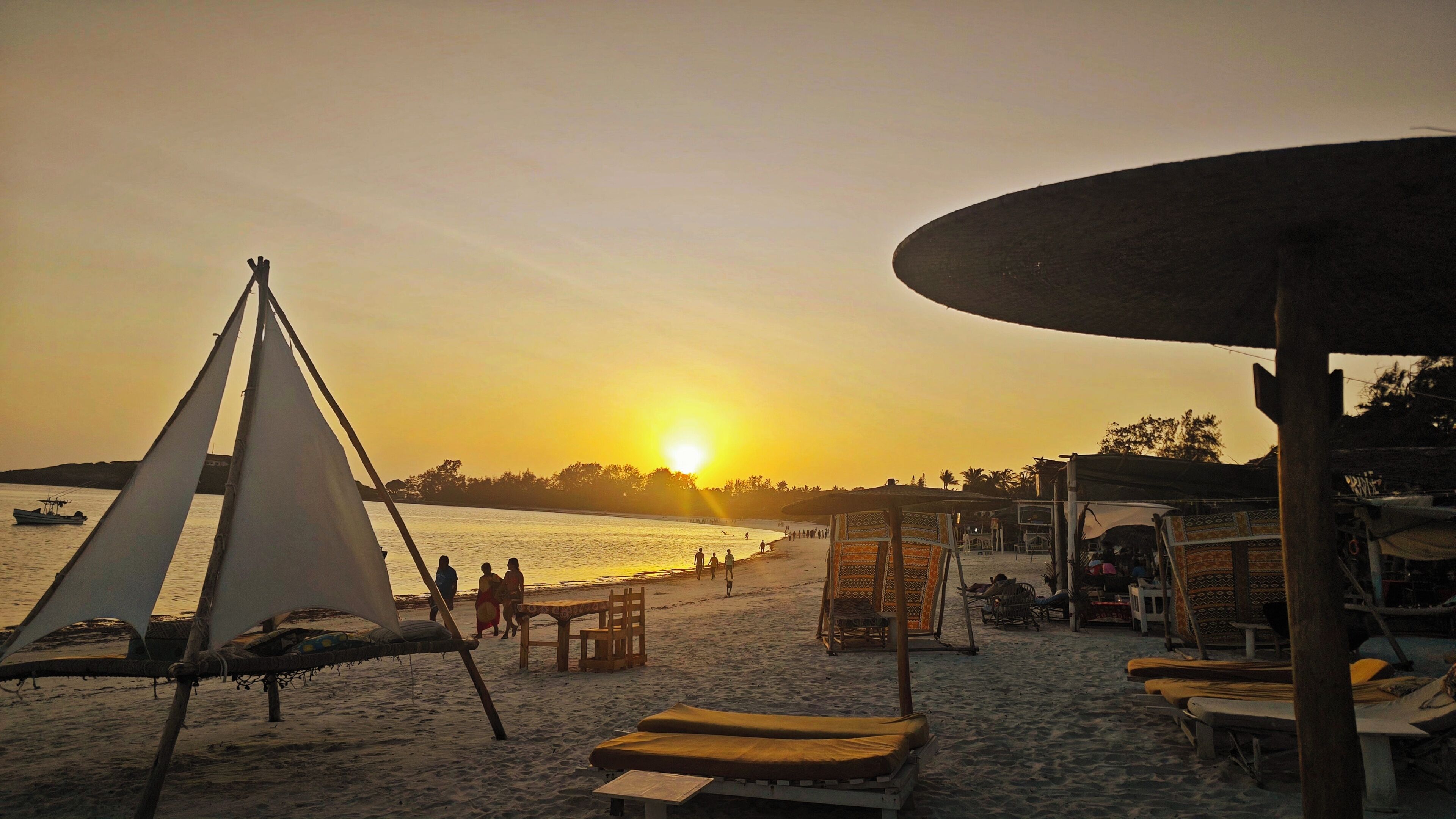 Uma praia com espreguiçadeiras, um veleiro e um pequeno pavilhão.
