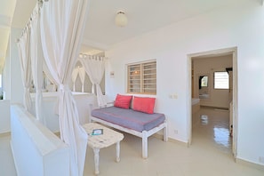 Quarto com cama, uma mesinha, janela com persianas e porta que dá para outro cômodo.