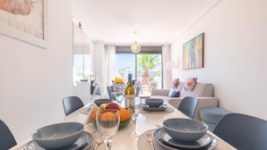 Uma área de refeições moderna com uma mesa branca posta para seis pessoas, com tigelas e pratos azul-escuros, uma garrafa de vinho e uma fruteira.