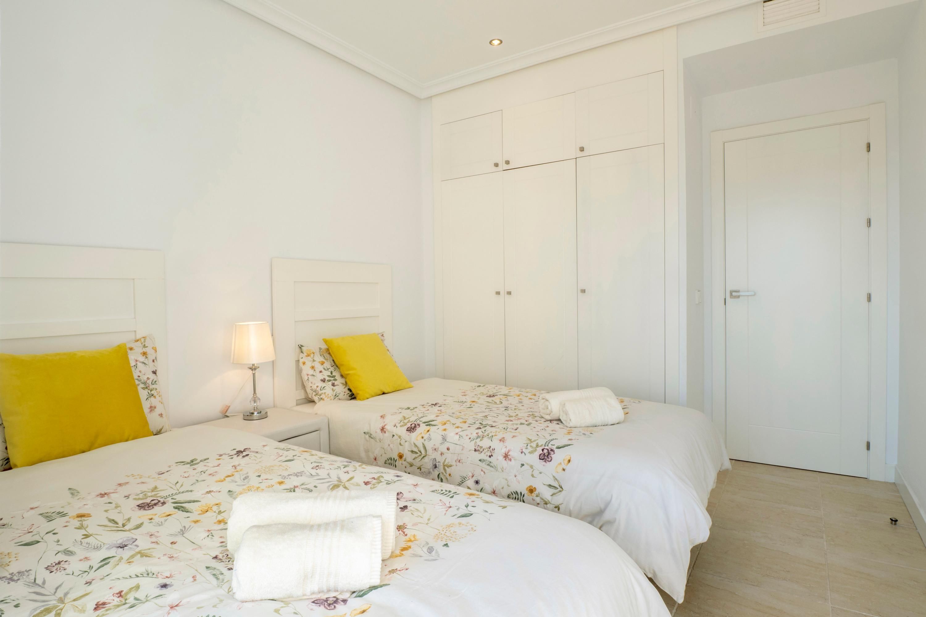 Quarto de hotel com duas camas, cada uma com roupa de cama florida e uma toalha branca. Há uma mesa de cabeceira com abajur, e um guarda-roupa com espelho.