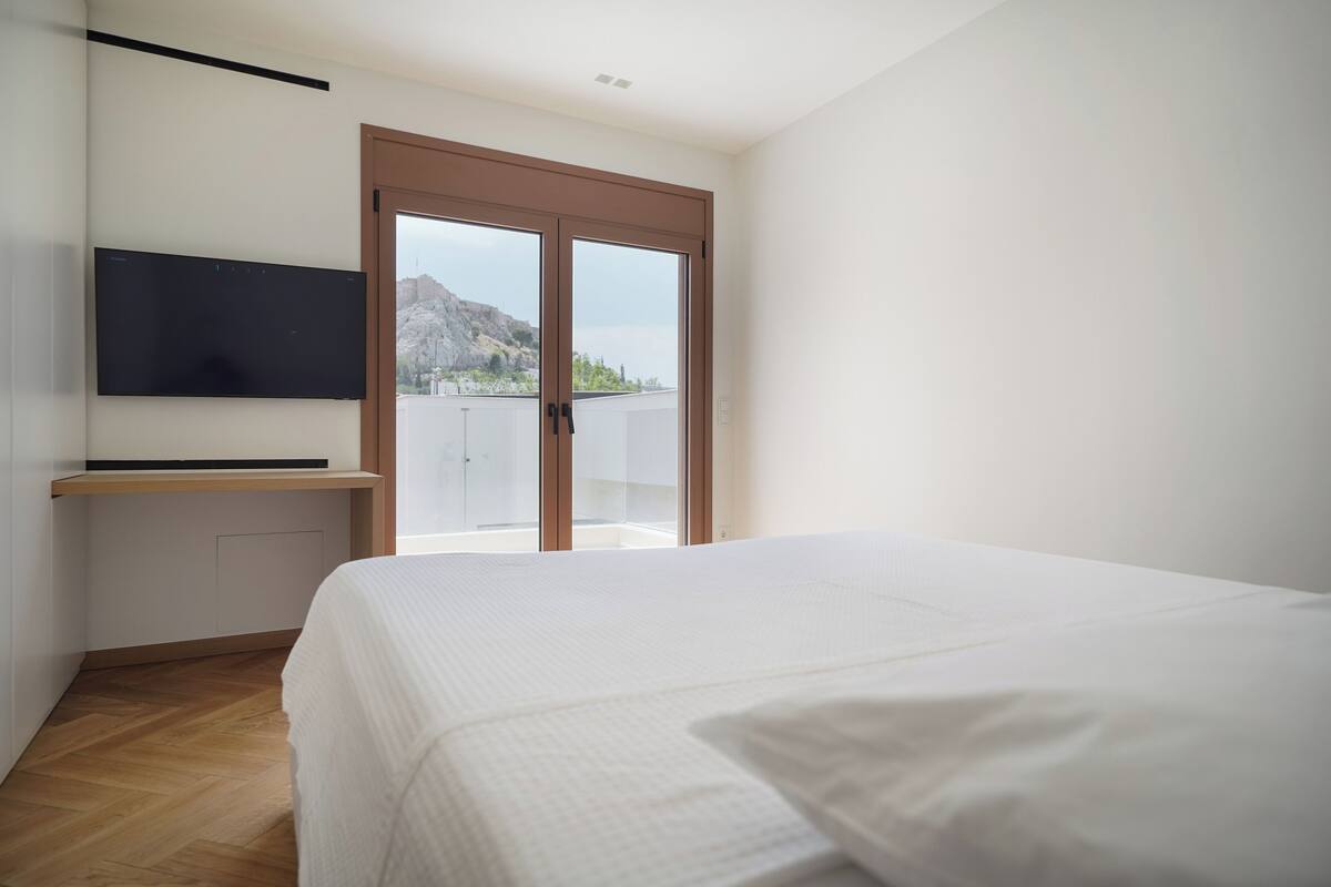 Quarto com cama, TV fixada na parede e porta de vidro deslizante que dá para uma varanda com vista para uma montanha.