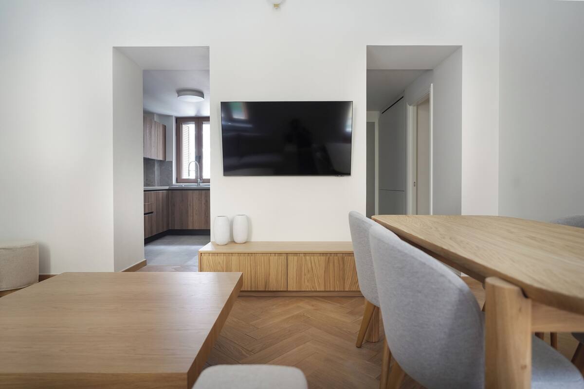 Uma sala de jantar moderna com mesa e cadeiras de madeira, uma TV de tela plana na parede e uma cozinha visível por uma porta aberta.