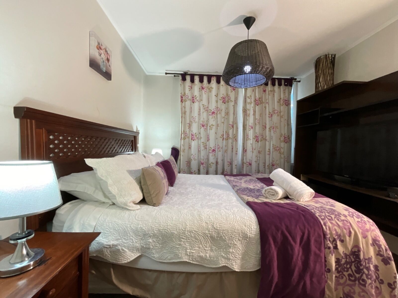 Quarto de hotel com cama, mesa de cabeceira com luminária, ventoinha no teto, televisão e cortinas florais.