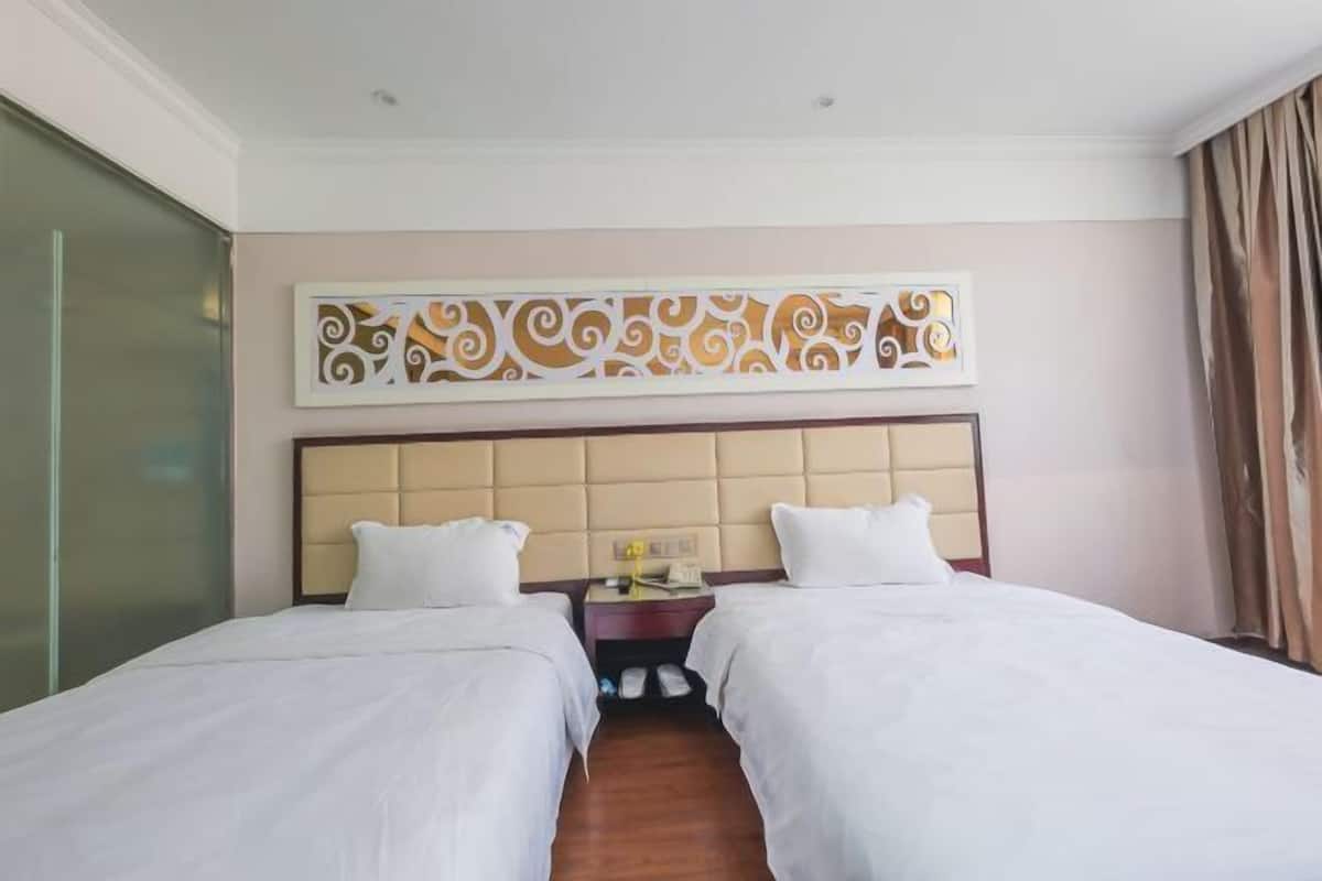 Quarto de hotel com duas camas, uma cabeceira decorativa, mesa de cabeceira com abajur e divisória de vidro.