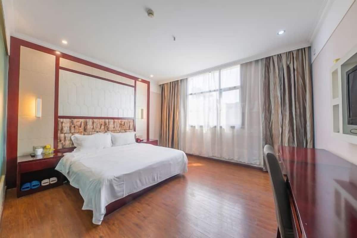 Quarto de hotel com uma cama grande, piso de madeira, uma escrivaninha e uma janela ampla com cortinas.