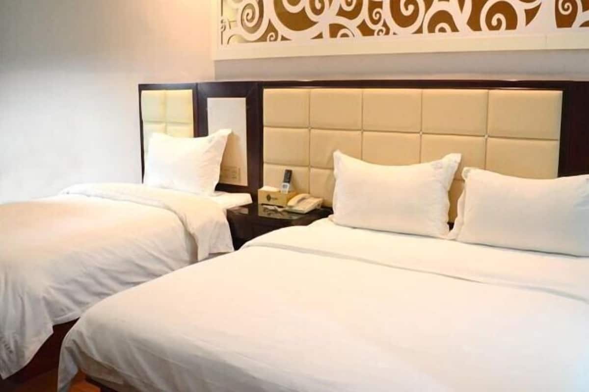 Quarto de hotel com duas camas, cabeceira com painel claro, mesa de cabeceira com telefone e luminária, e painel decorativo na parede.