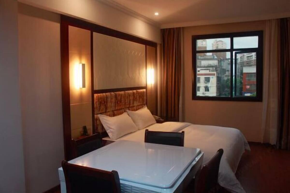 Quarto de hotel com cama, mesa de cabeceira, cadeira, janela com vista para edifícios e espelho fixado na parede.