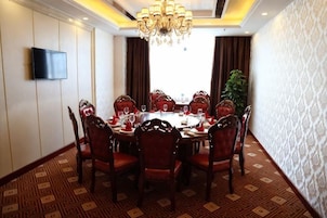 Uma sala de conferências com uma mesa redonda, cadeiras estofadas em vermelho, um candelabro e uma televisão na parede.