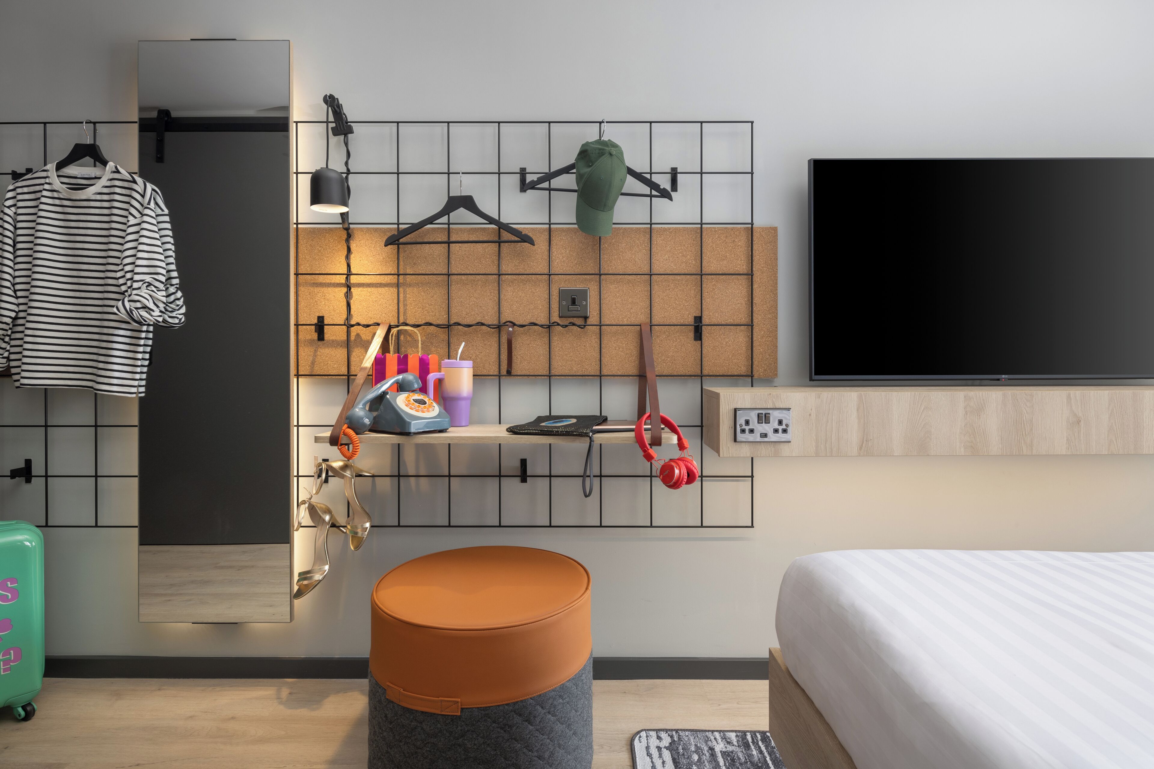 Quarto de hotel moderno com cama, televisão de tela plana grande, guarda-roupa, espelho, pequeno espaço de estar e prateleira na parede com objetos pessoais.