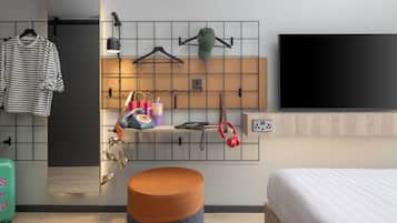 Quarto de hotel moderno com cama, televisão de tela plana grande, guarda-roupa, espelho, pequeno espaço de estar e prateleira na parede com objetos pessoais.