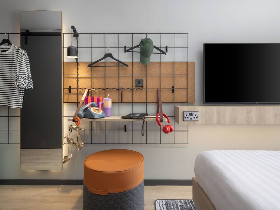 Quarto de hotel moderno com cama, televisão de tela plana grande, guarda-roupa, espelho, pequeno espaço de estar e prateleira na parede com objetos pessoais.