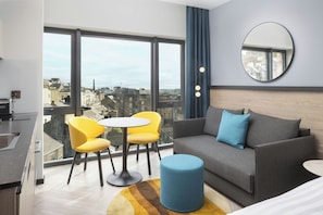 Quarto de hotel moderno com varanda, sofá, duas cadeiras amarelas, mesa redonda, banquinho azul e um espelho grande.
