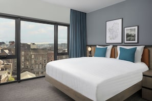 Quarto de hotel com uma cama grande, vista para a cidade, mesa de cabeceira e duas fotos emolduradas na parede.