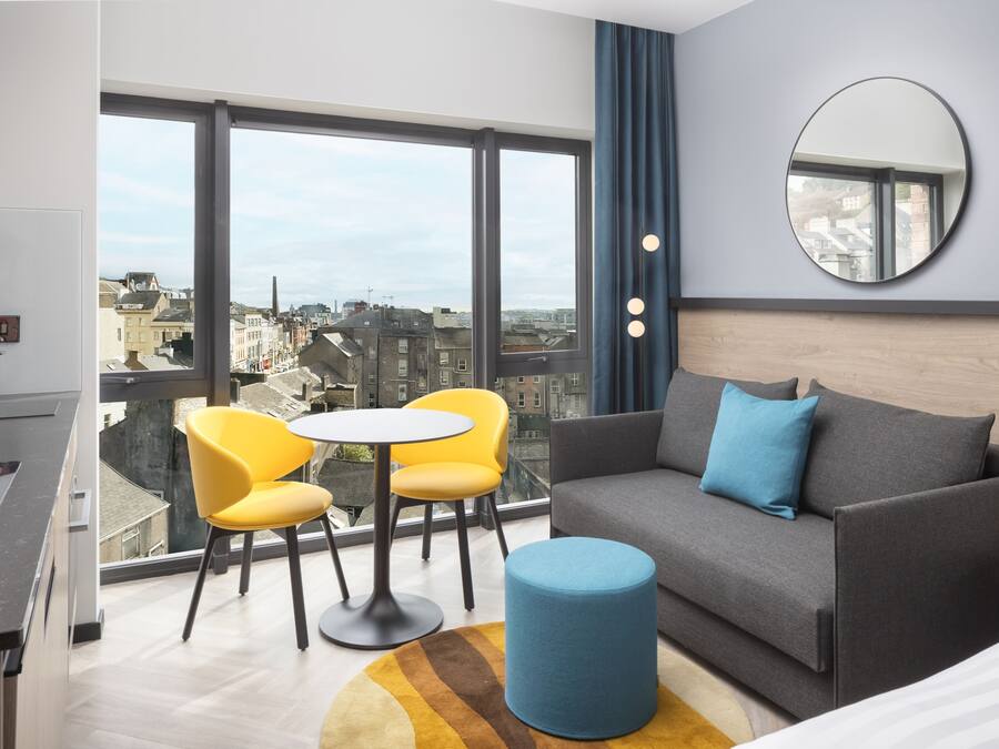 Quarto moderno com varanda, um sofá cinzento, cadeiras amarelas, um espelho redondo e vista para a cidade.