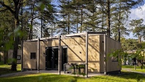 Uma cabana moderna de um andar em madeira, com porta de vidro, um pequeno alpendre com mesa e cadeiras, e arredores com relvado e árvores.