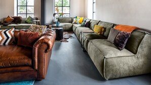 Uma sala de estar moderna com sofás de couro, uma mesa de centro e um tapete. Há janelas que proporcionam luz natural e vista para o verde lá fora.