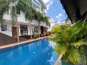 Uma piscina com borda azulejada, cercada por palmeiras e um edifício com várias janelas e uma escadaria.