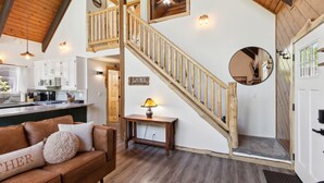 Uma sala de estar ampla com escada de madeira, um sofá marrom aconchegante, uma cozinha ao fundo e uma placa com a inscrição 