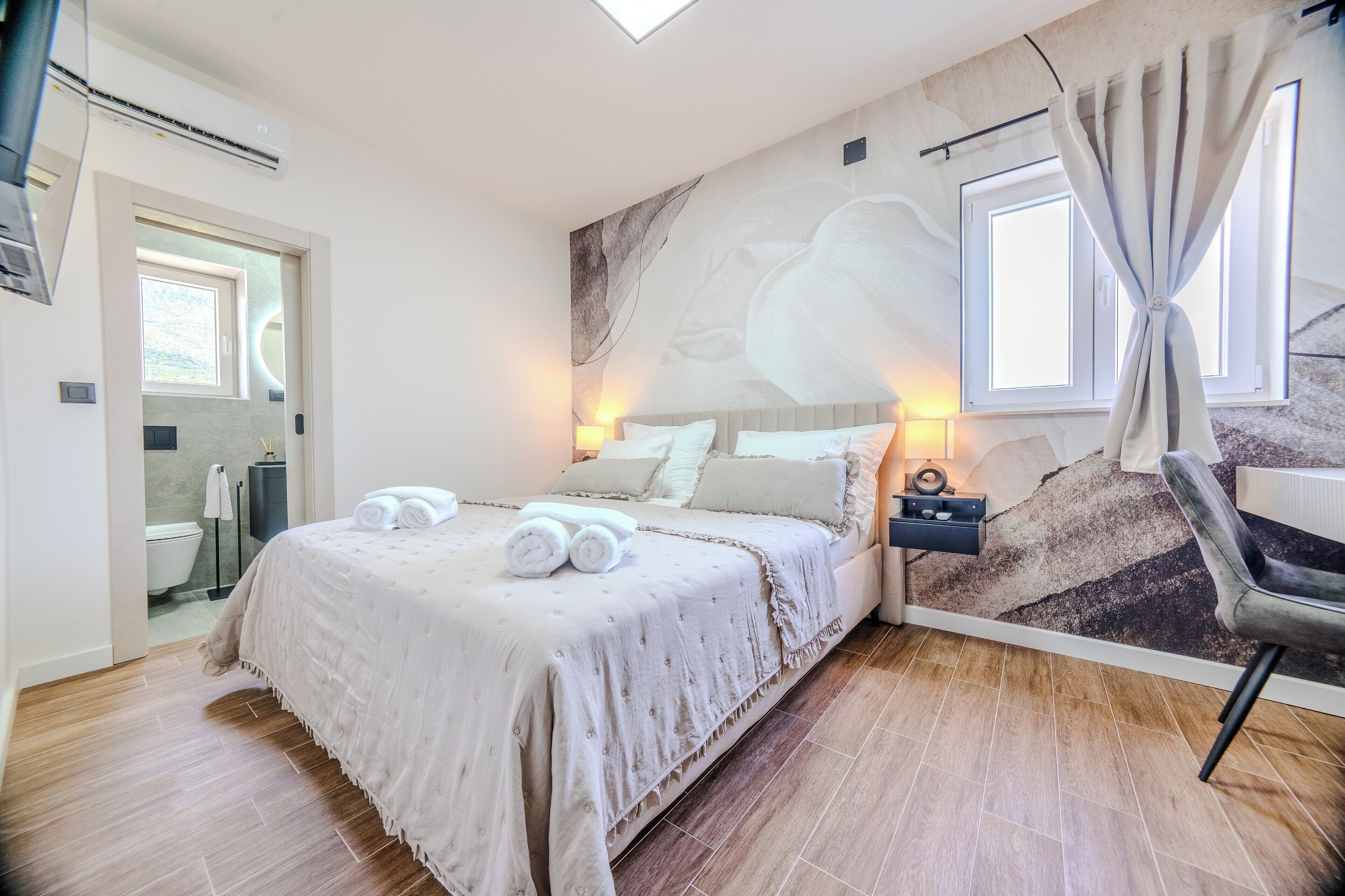 Quarto com cama, mesa de cabeceira, luminária na parede, janela com cortinas e banheiro visível por uma porta aberta.