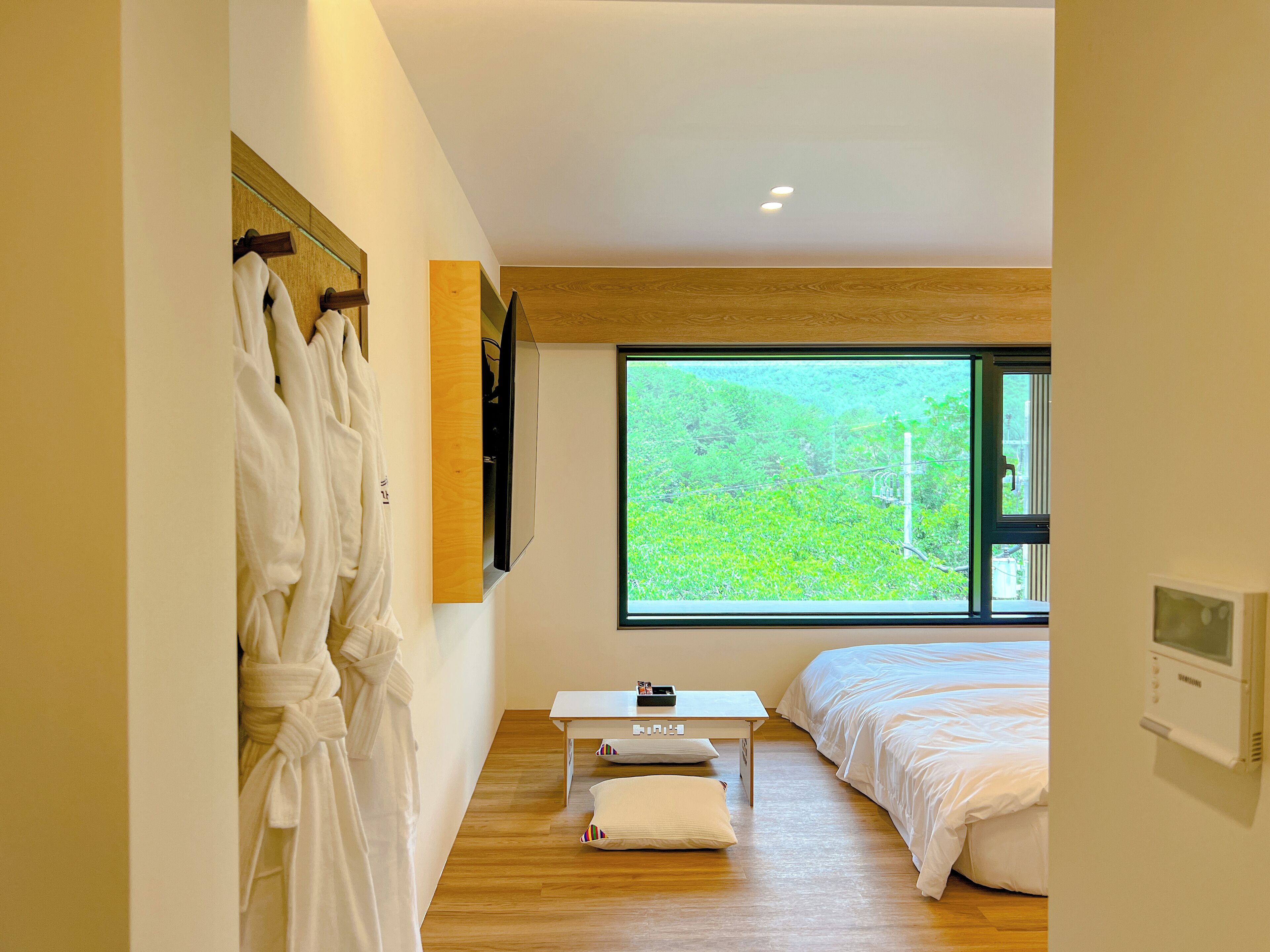 Quarto moderno com uma grande janela, uma cama, uma mesa de cabeceira com abajur e um roupão pendurado na parede.