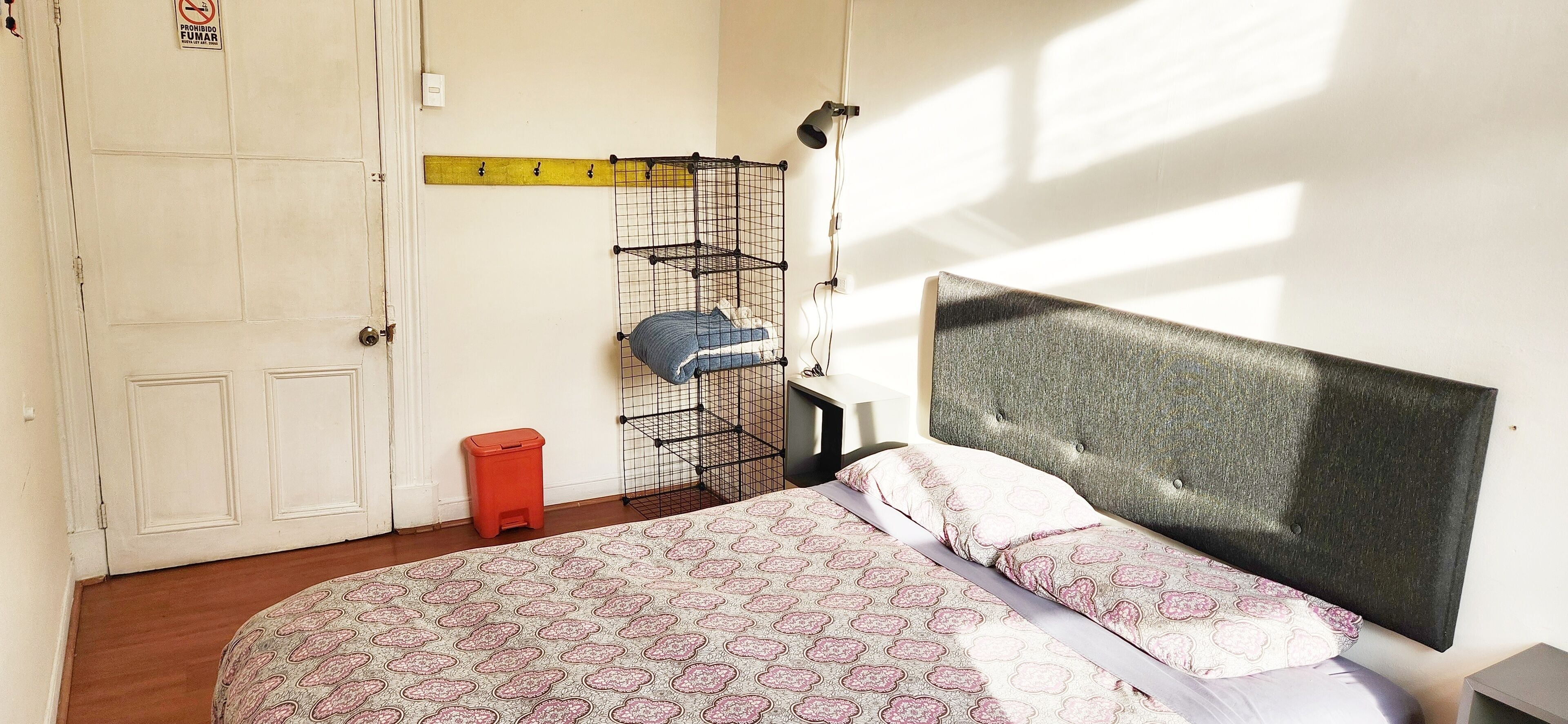 Quarto com cama, guarda-roupa, um pequeno banquinho, prateleira com roupas e luminária na parede.
