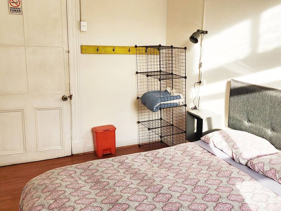 Quarto com cama, guarda-roupa, um pequeno banquinho, prateleira com roupas e luminária na parede.
