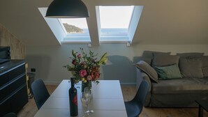 Uma área de jantar com mesa, cadeiras, um vaso de flores, uma garrafa de vinho e uma luminária pendente. Há duas claraboias acima da mesa, e um sofá é visível ao fundo.