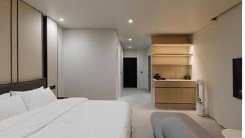 Quarto de hotel moderno com uma cama grande, mesa de cabeceira, sofá, televisão, minibar e banheiro visível ao fundo.