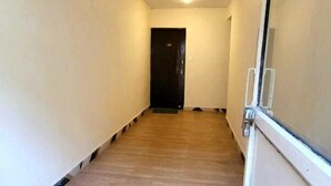 Um corredor estreito com soalho de madeira, paredes brancas e uma porta preta no final.