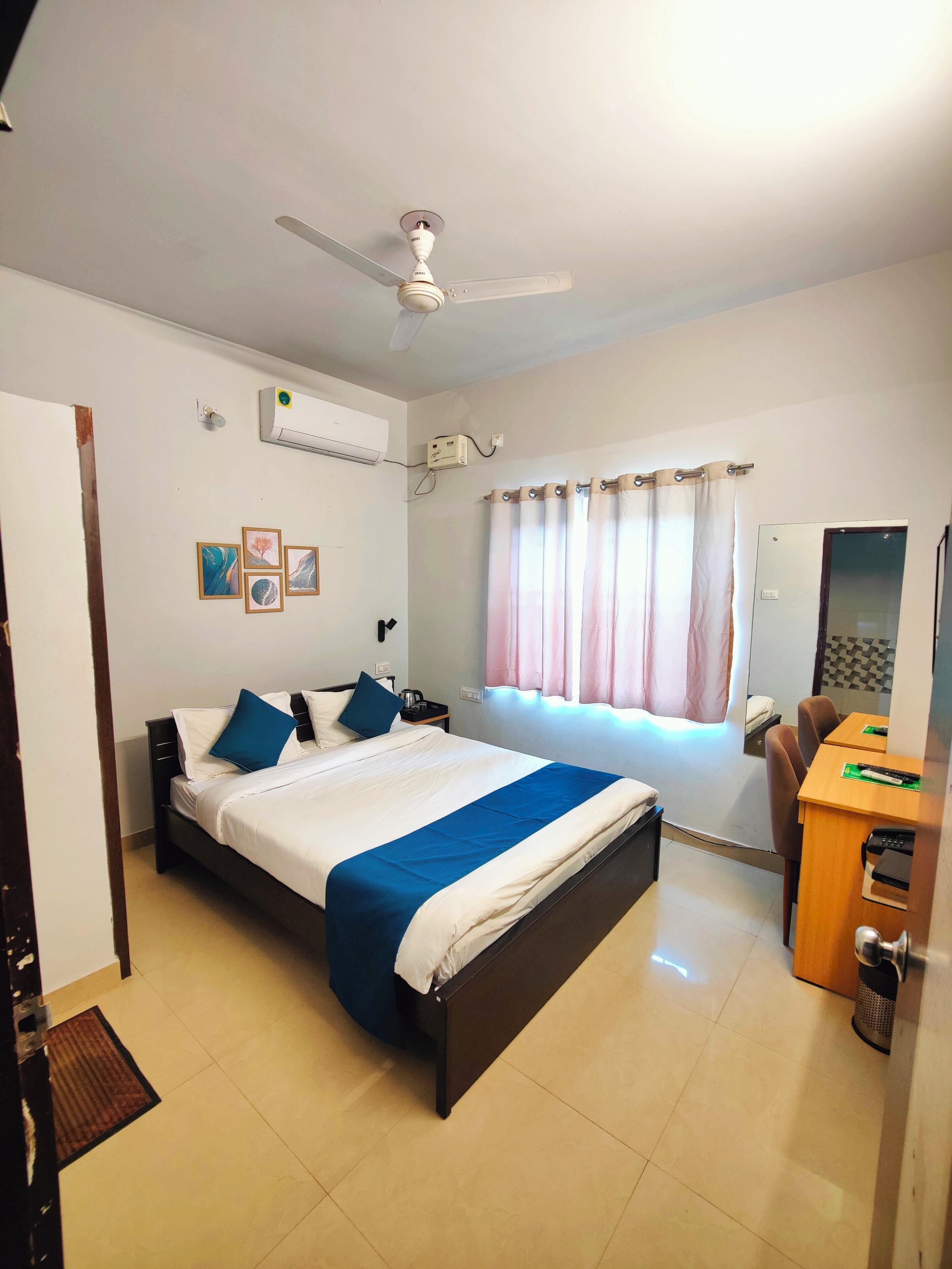 Quarto de hotel com cama, escrivaninha, cadeira, ventoinha no teto e janela com cortinas.